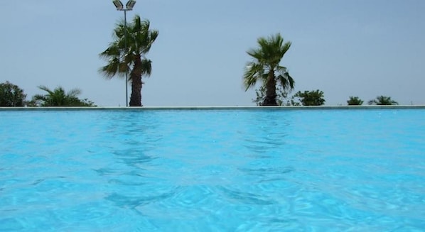 Piscina com água azul cristalina, rodeada por palmeiras e um céu claro.