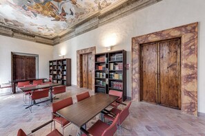 Uma biblioteca com uma longa mesa de madeira e cadeiras vermelhas, rodeada por estantes de livros e grandes portas de madeira.
