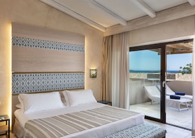 Quarto de hotel com cama, mesa de cabeceira, painel de cabeceira estampado, uma janela ampla com vista para o mar e varanda com cadeira e mesa.
