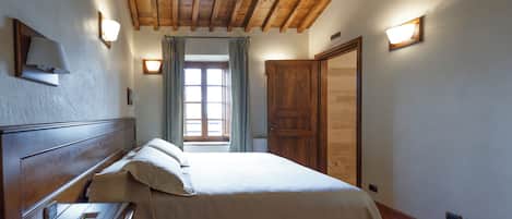 Quarto com teto de madeira, cama com roupa de cama branca, mesa de cabeceira, janela com cortinas e guarda-roupa de madeira.
