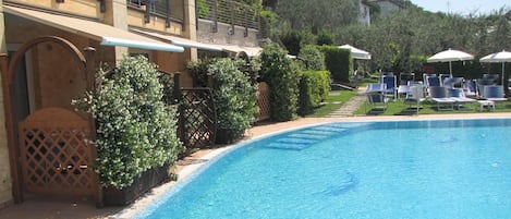 Piscina com espreguiçadeiras e guarda-sóis, cercada por vegetação e um edifício com varandas.