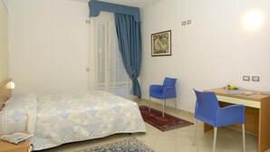 Quarto com uma cama, escrivaninha, duas cadeiras, janela com cortinas e luminária fixada na parede.