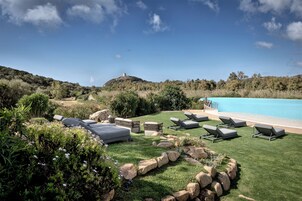 Uma área externa ajardinada com piscina, espreguiçadeiras e uma colina ao fundo.