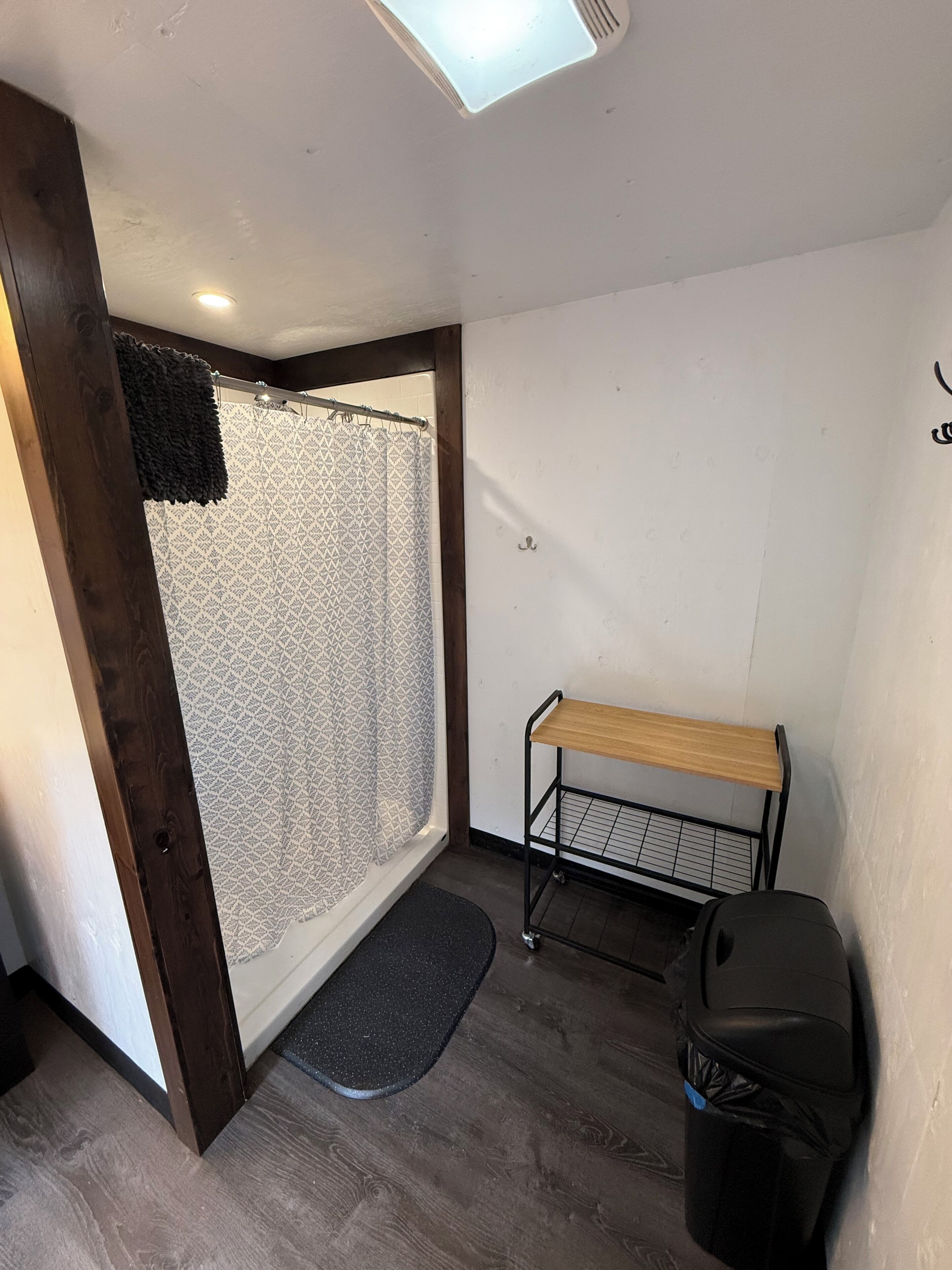 Um quarto pequeno e bem iluminado, com chuveiro, um banco, uma lixeira e um claraboia.