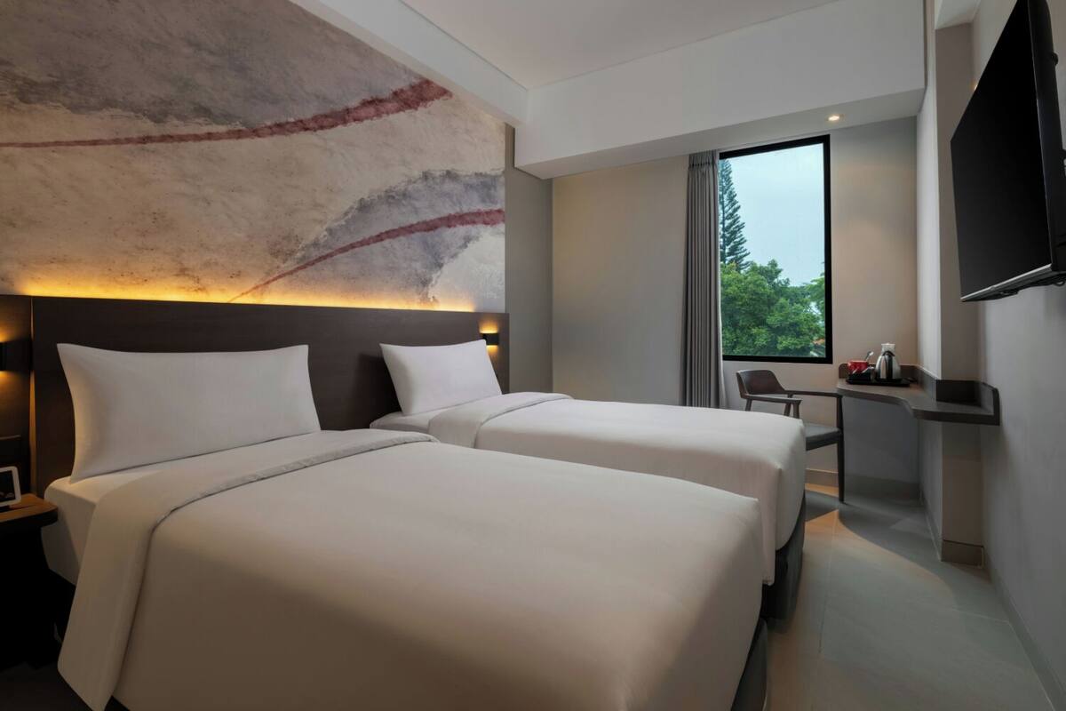 Quarto de hotel moderno com duas camas, uma escrivaninha e uma janela grande com vista para o verde.