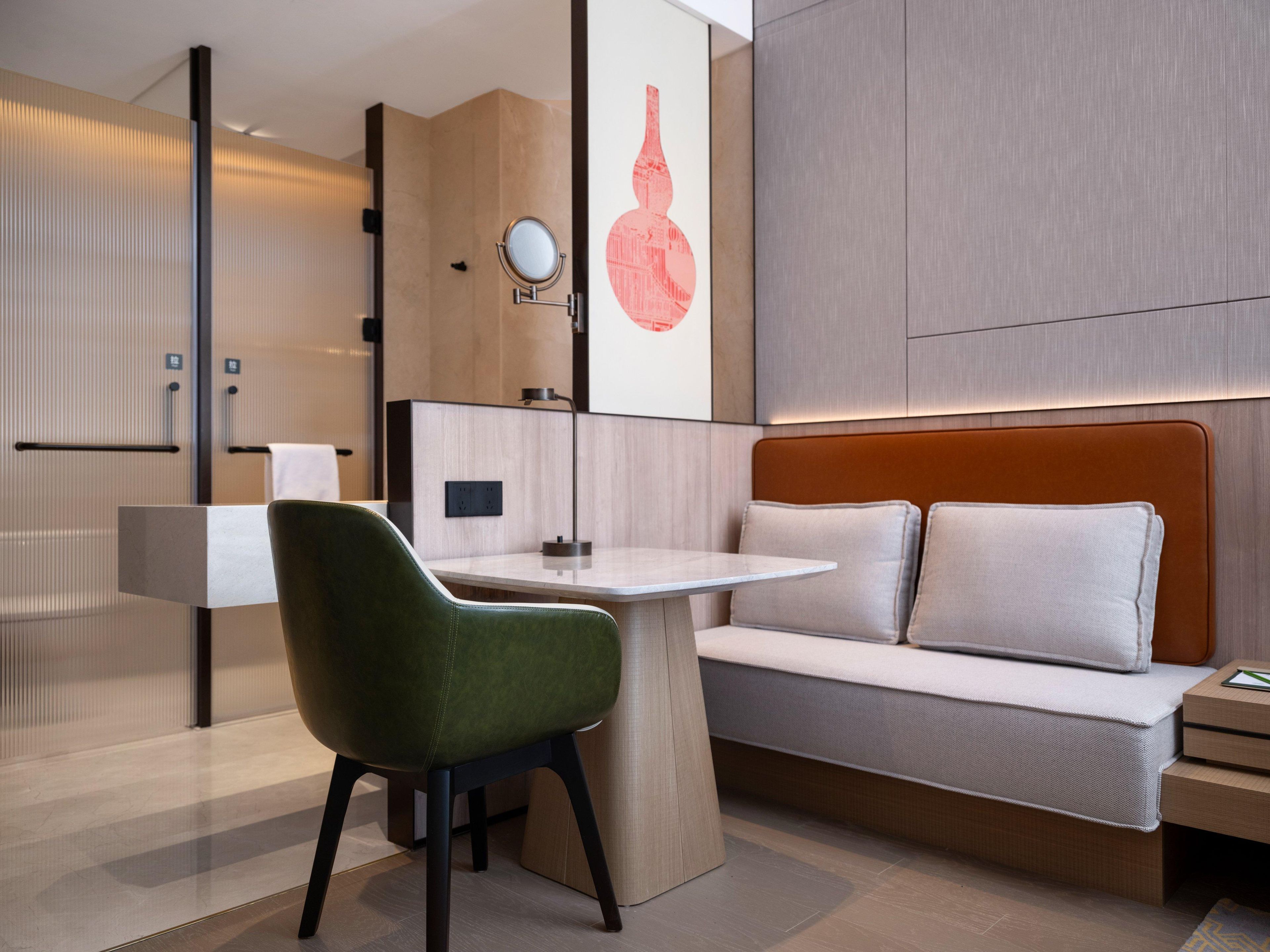 Quarto de hotel moderno com uma cadeira verde, uma mesa redonda, um sofá com almofadas e um espelho fixado na parede.