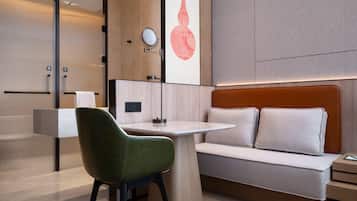 Quarto de hotel moderno com uma cadeira verde, uma mesa redonda, um sofá com almofadas e um espelho fixado na parede.