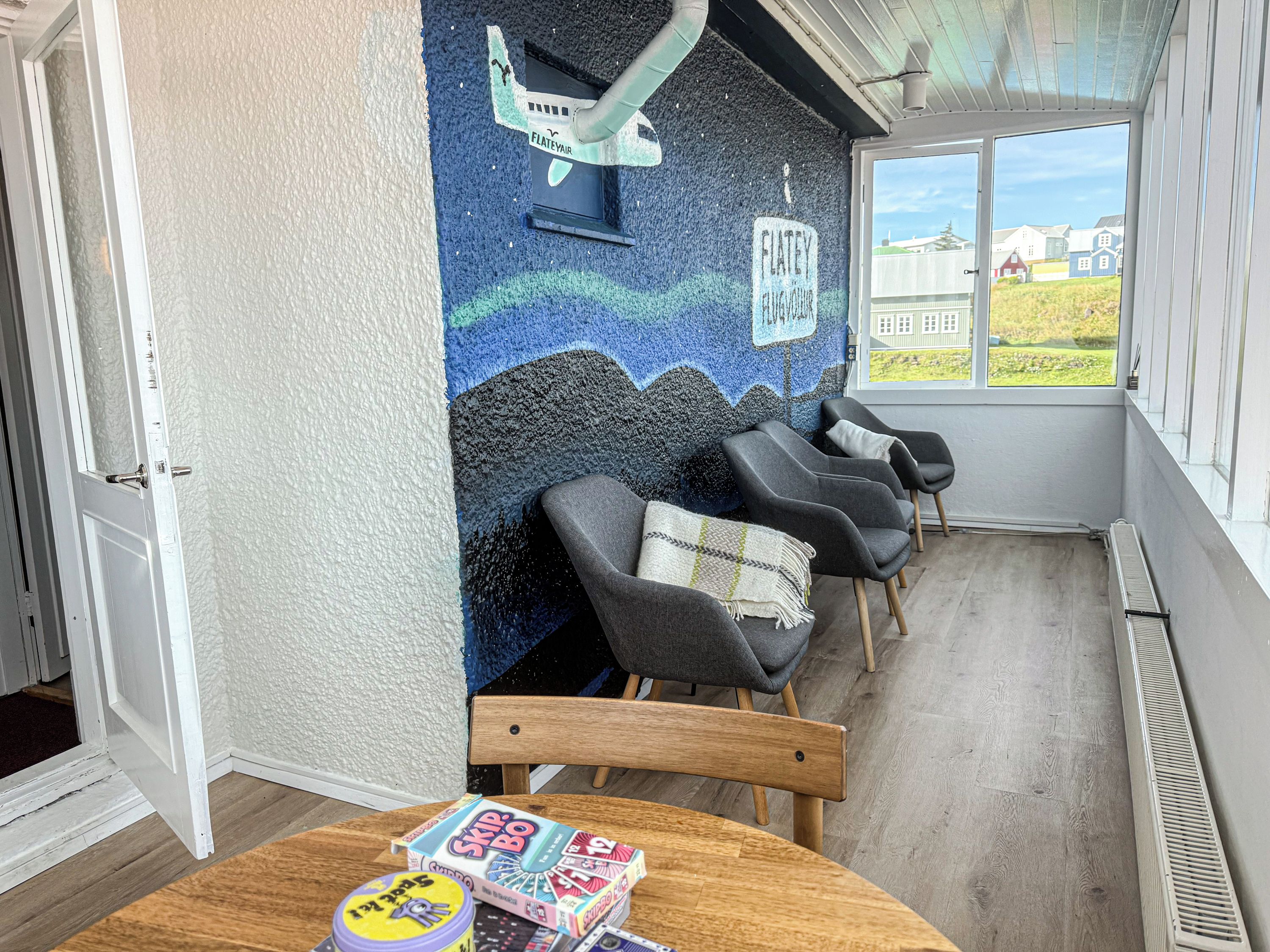 Quarto com parede azul texturizada, uma mesa de madeira com um brinquedo de barco Skipper e vista para casas pela janela.