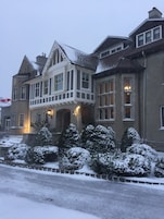 Uma casa grande, de vários andares, com uma varanda frontal imponente e paisagismo coberto de neve.
