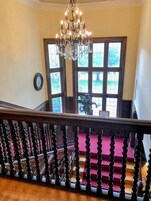 Uma escadaria imponente com um candelabro, um relógio e vista para o exterior através de amplas janelas.