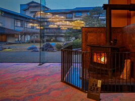 Um quarto em estilo japonês tradicional, com piso de esteira de tatame, um fogão a lenha e vista para uma área residencial com edifícios tradicionais.