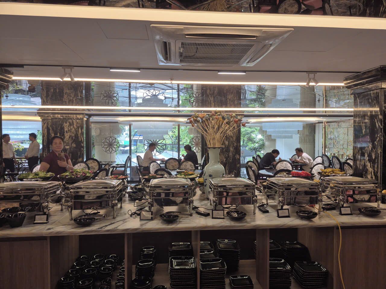 Disposição de buffet com várias estações de serviço, um vaso grande com plantas secas e vista para a rua.