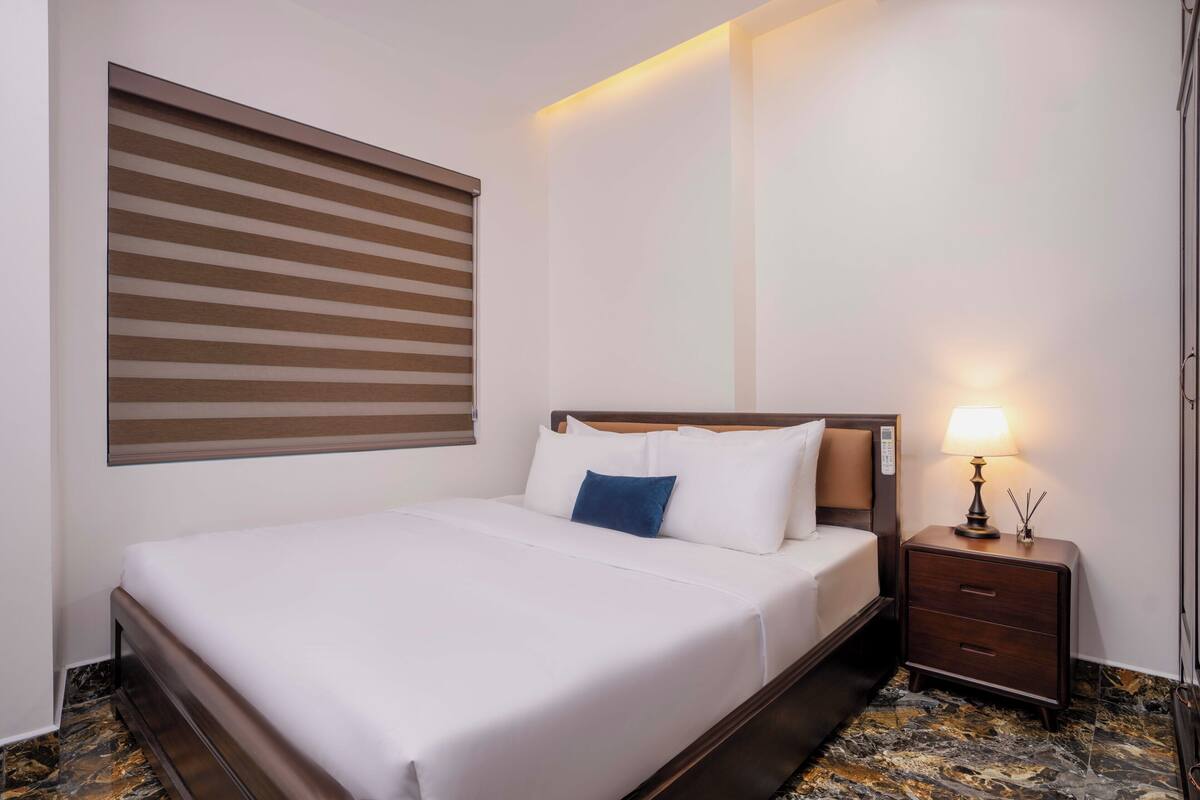 Quarto de hotel com cama, mesa de cabeceira com luminária, janela com persianas e piso de mármore.