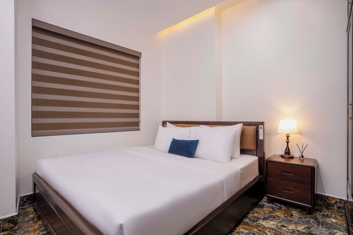 Quarto de hotel com uma cama, uma mesa de cabeceira com abajur e uma janela com persianas.