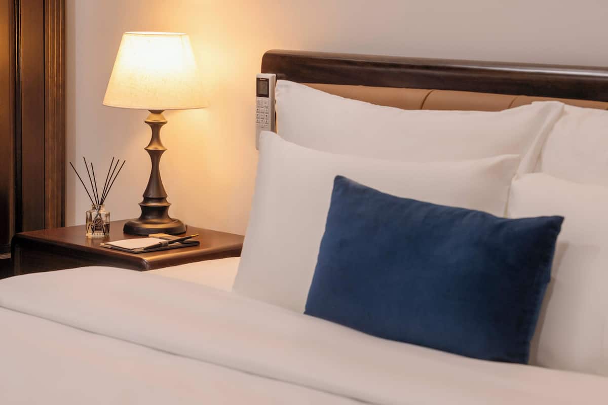 Cama bem arrumada com almofadas brancas e azuis, luminária de cabeceira e pequena mesa com caneta e um recipiente de vidro.