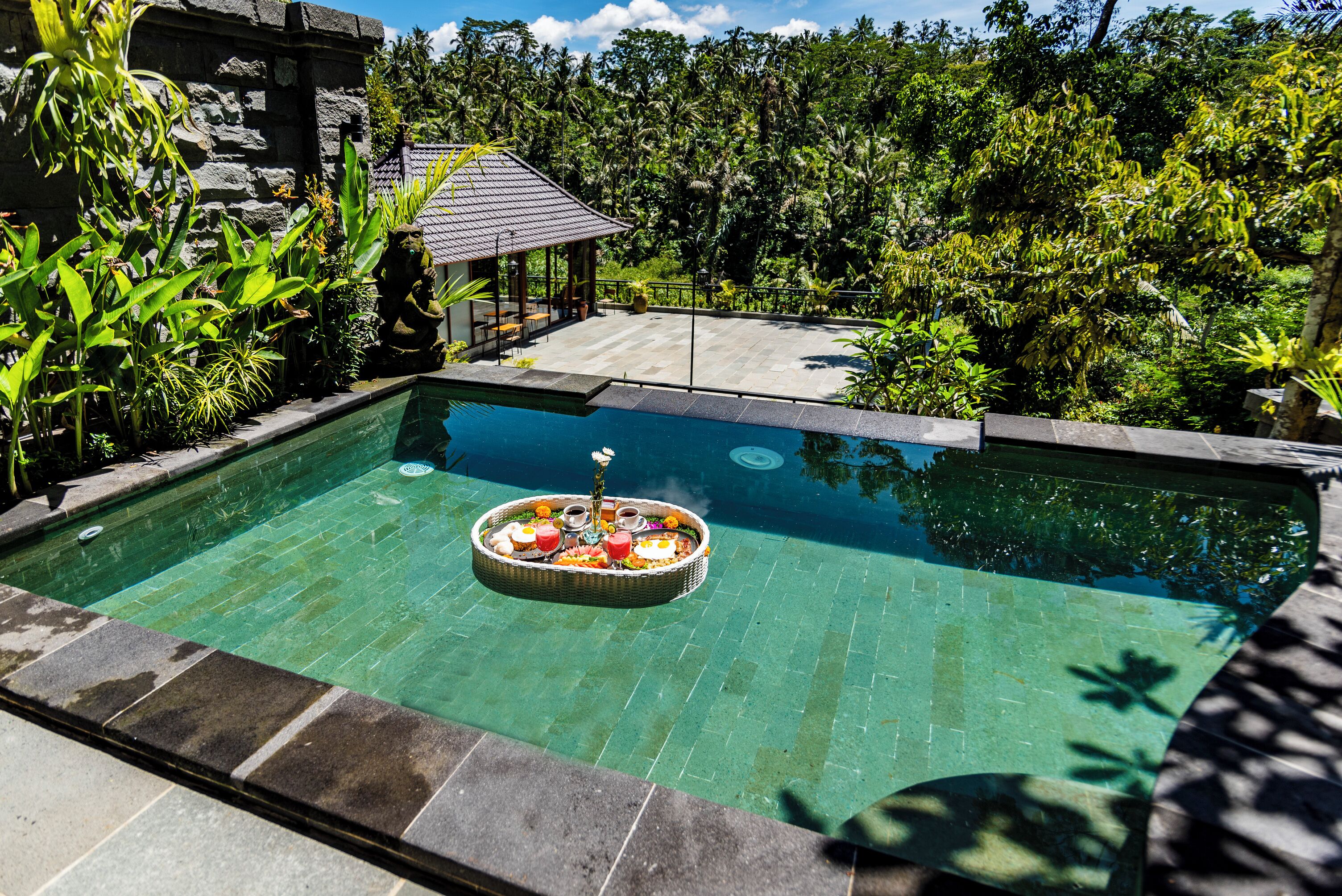 Uma piscina com área de bar flutuante, rodeada por vegetação exuberante e estruturas de pedra.