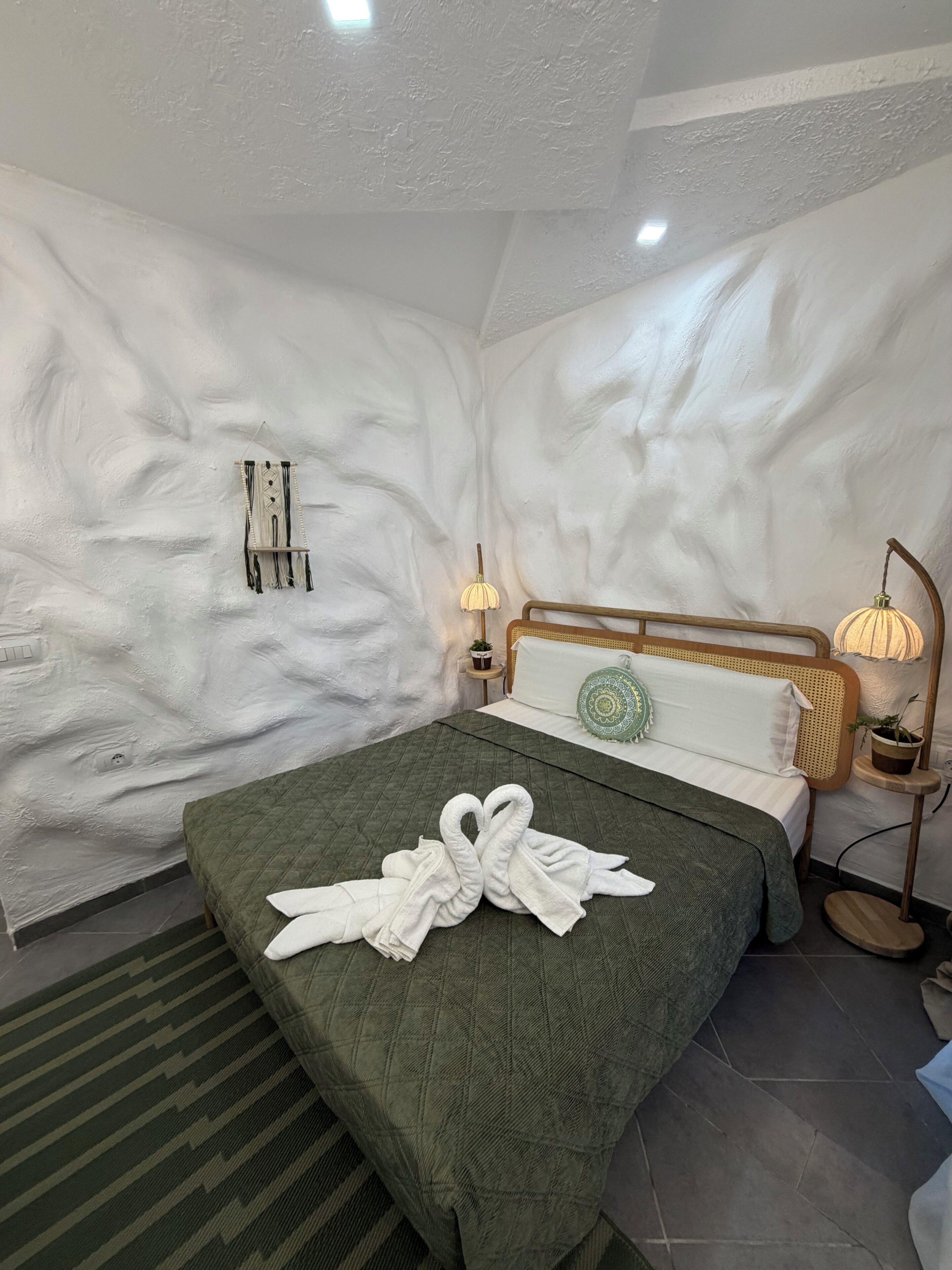 Quarto com cama, uma tapeçaria de macramé na parede, luminária de chão e mesa de cabeceira com planta.