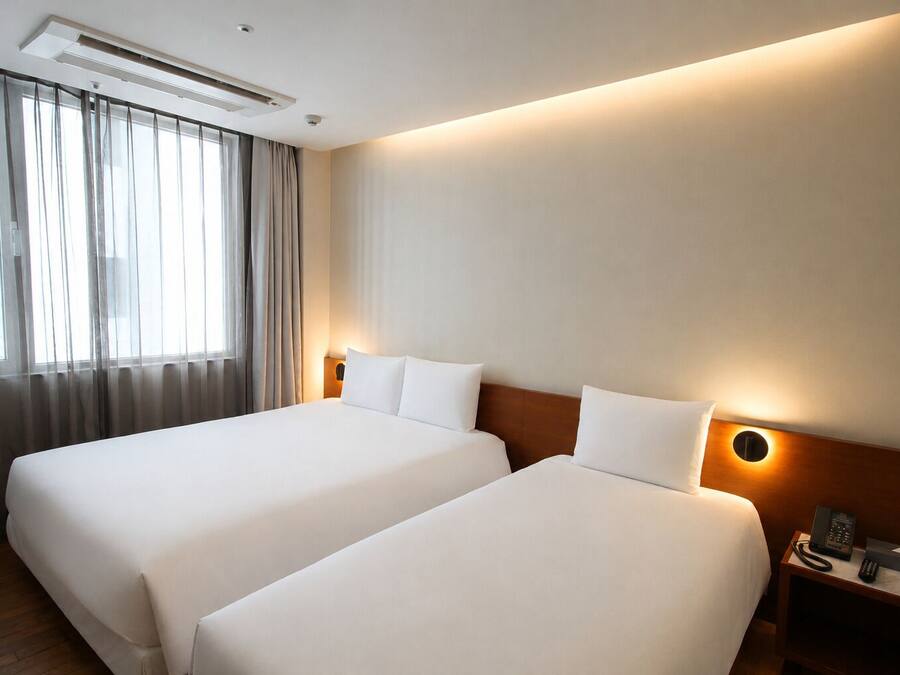 Quarto de hotel com duas camas, uma mesa de cabeceira com luminária e uma janela com cortinas.