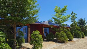 Uma casa de dois andares com fachada azul e vermelha, cercada por vegetação bem cuidada e um caminho de acesso pavimentado.