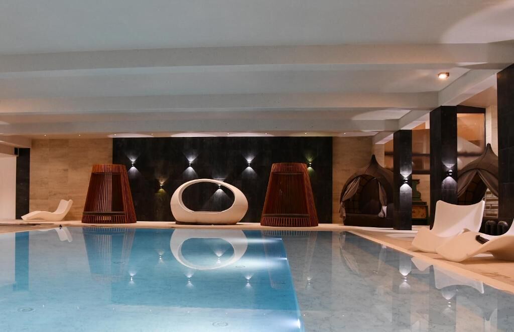 Uma área interna de piscina moderna, com piscina transparente, espreguiçadeiras e arranjos de assentos originais.