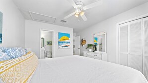 Quarto com uma cama grande, um ventilador de teto, um quadro de uma cena de praia, um espelho e um guarda-roupa.