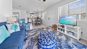 Uma sala de estar ampla, com um sofá azul, um banquinho e uma televisão grande sobre um móvel branco.
