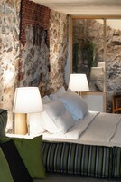 Quarto com parede de pedra, cama com roupa de cama branca, duas luminárias de mesa e vista para o exterior através de uma porta de vidro.
