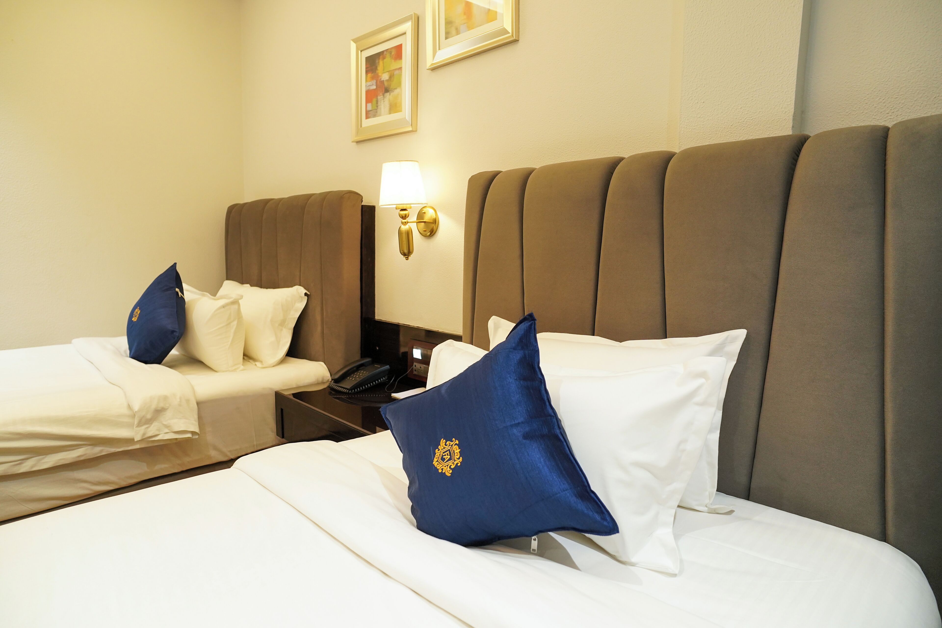 Quarto de hotel com duas camas, cada uma com roupa de cama branca e almofadas azuis, uma mesa de cabeceira com abajur e telefone.