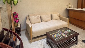 Uma sala de estar com um sofá bege, uma mesa de centro com contas coloridas, uma cadeira de madeira, uma planta e quatro relógios na parede.