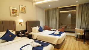Quarto de hotel com duas camas, uma mesa, uma cadeira e vista para a paisagem urbana através de grandes janelas.