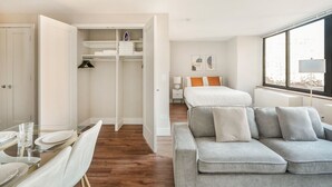 Quarto moderno e bem iluminado, com área de refeições, um sofá cinza com almofadas e uma cama com travesseiros laranja, além de um armário com prateleiras.