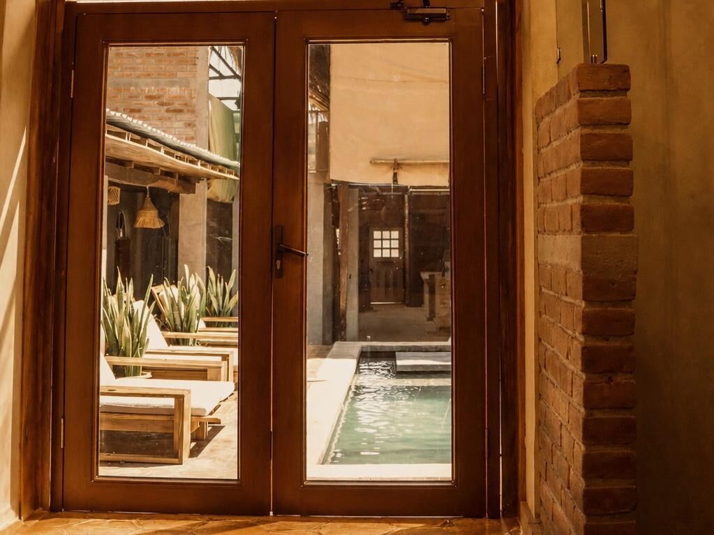Uma porta de madeira dá acesso a uma área de piscina com espreguiçadeiras e um muro de tijolos.