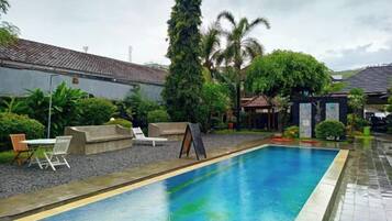 Piscina com espreguiçadeiras e uma pequena mesa, rodeada por vegetação e um edifício com telhado de azulejos.