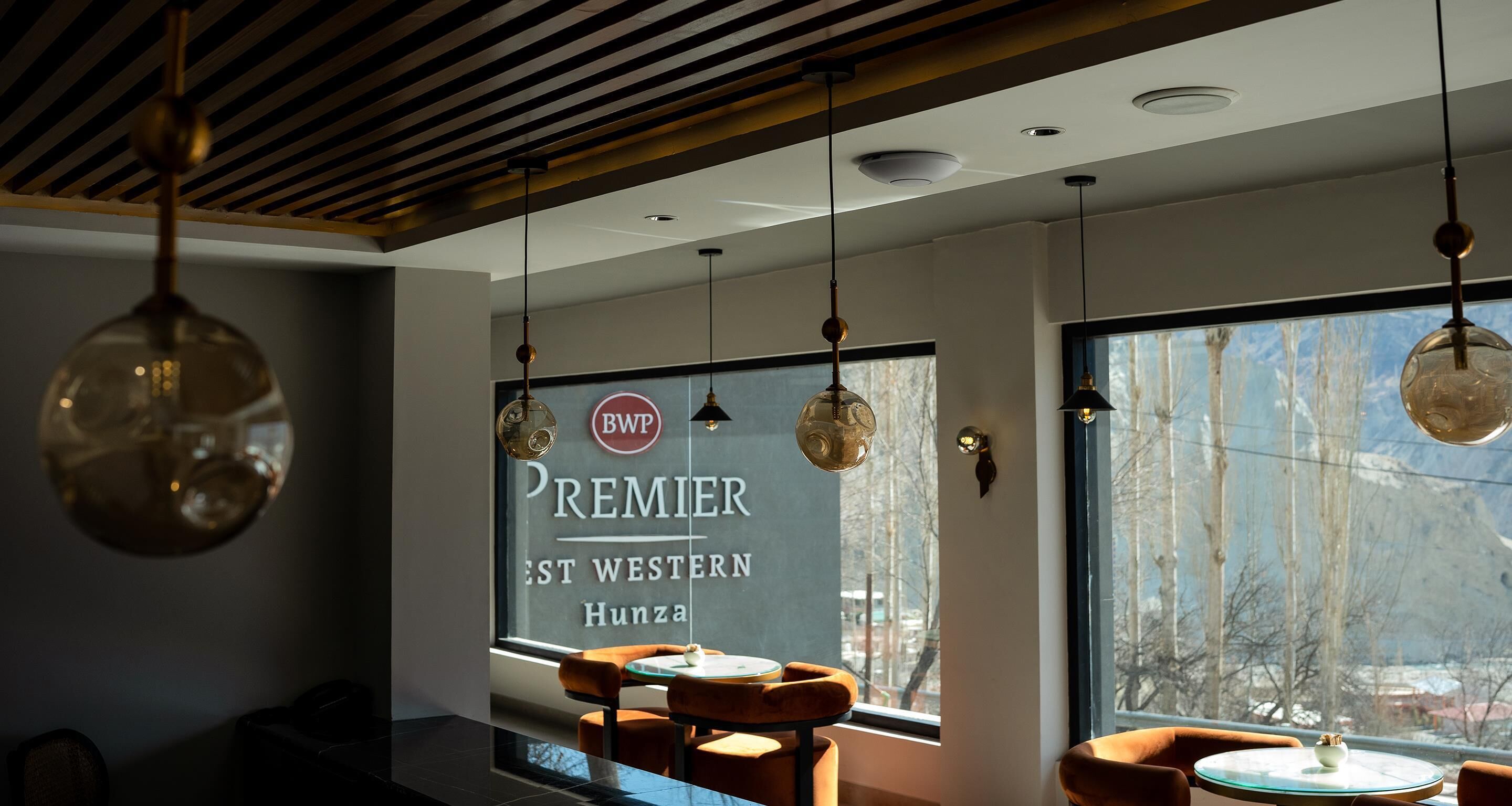 Um interior moderno com luminárias pendentes, uma janela grande com vista para montanhas e uma placa com a inscrição 'Premier West Western Hunza'.