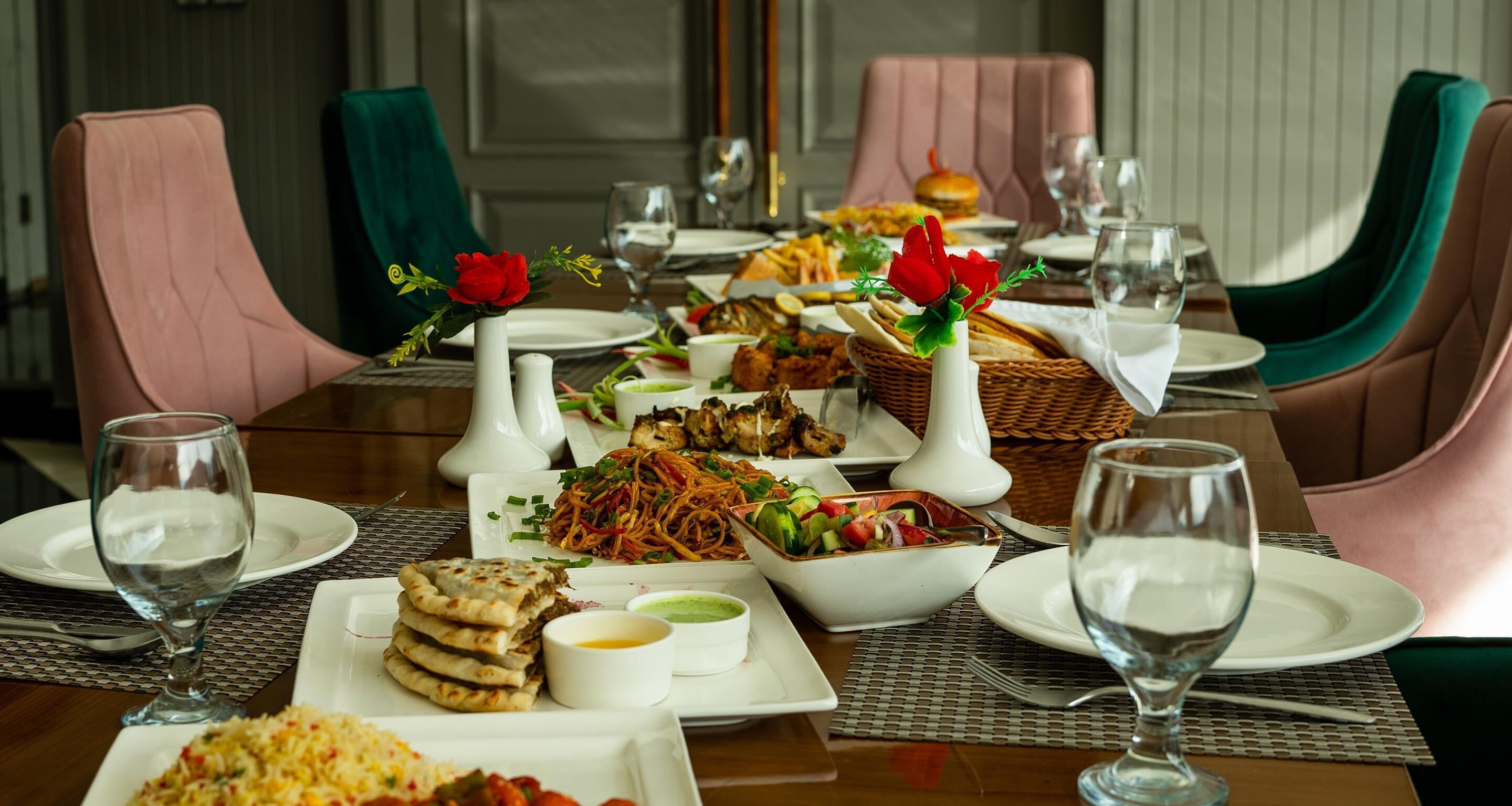 Uma mesa de jantar posta com diversos pratos, incluindo salada, carnes grelhadas e pães achatados, em um restaurante com cadeiras rosa e azul-turquesa.