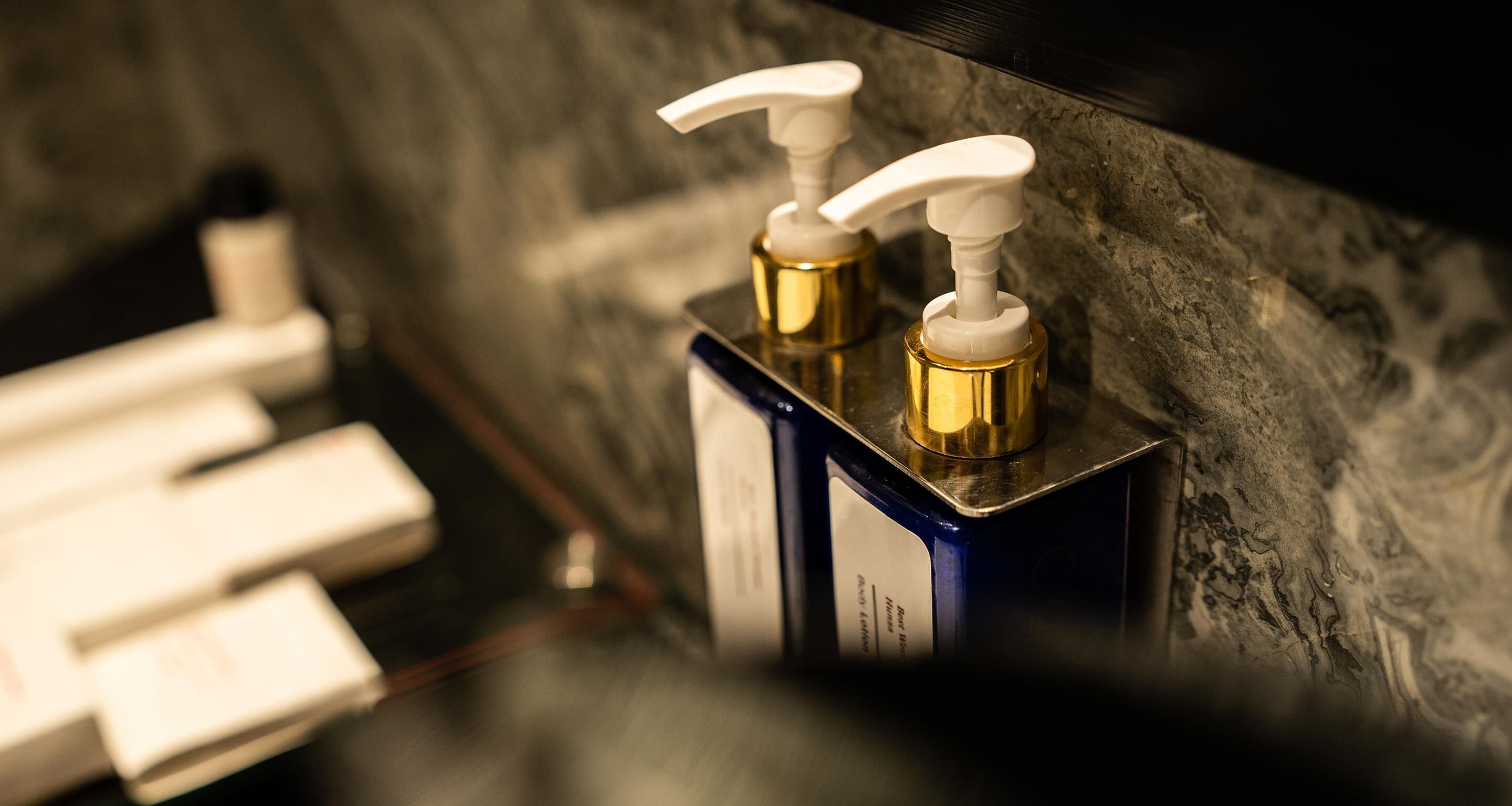 Casa de banho com parede de mármore, dosador de sabonete dourado e azul, e frasco branco.
