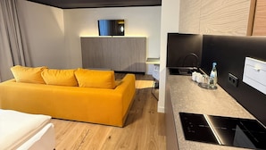 Quarto de hotel moderno com sofá amarelo, piso de madeira, cozinha planejada com fogão e uma janela grande.