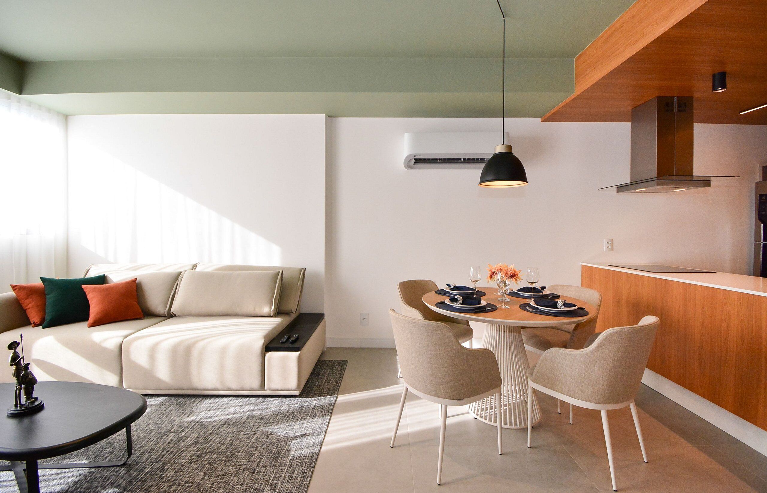 Uma sala de estar moderna com sofá, mesa de jantar e cadeiras. Há uma cozinha com bancada e luminária pendente.
