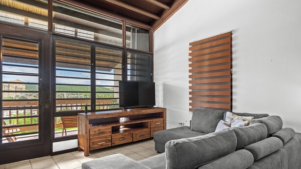 Uma sala de estar com um sofá cinza, um móvel de televisão em madeira e uma janela grande com persianas.
