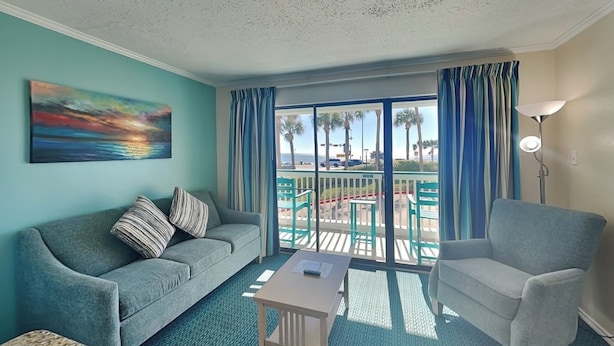 Quarto de hotel com carpete azul, sofá, cadeira, mesa pequena e um quadro na parede.