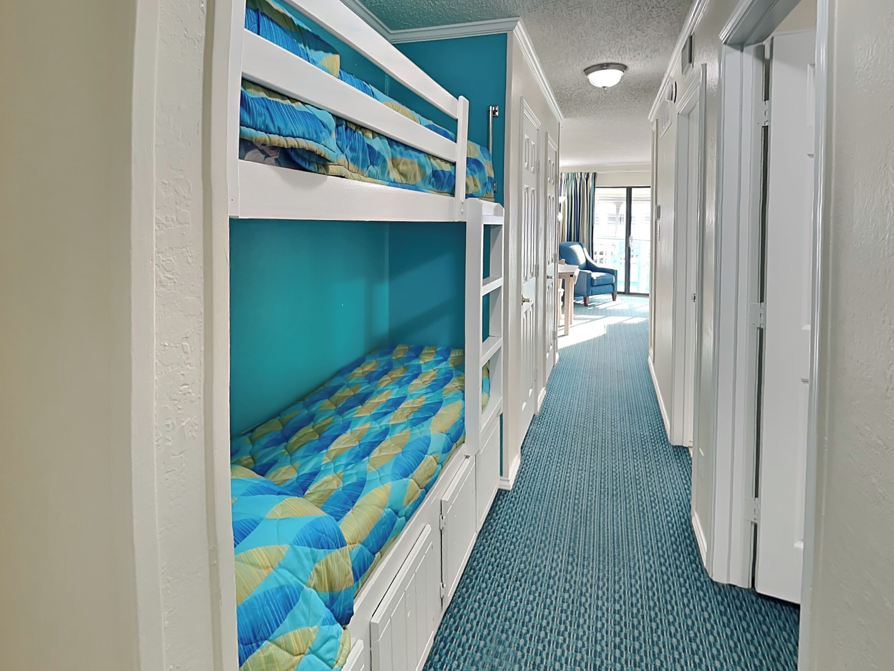 Um corredor com beliches, uma cadeira e uma porta que dá para um quarto.