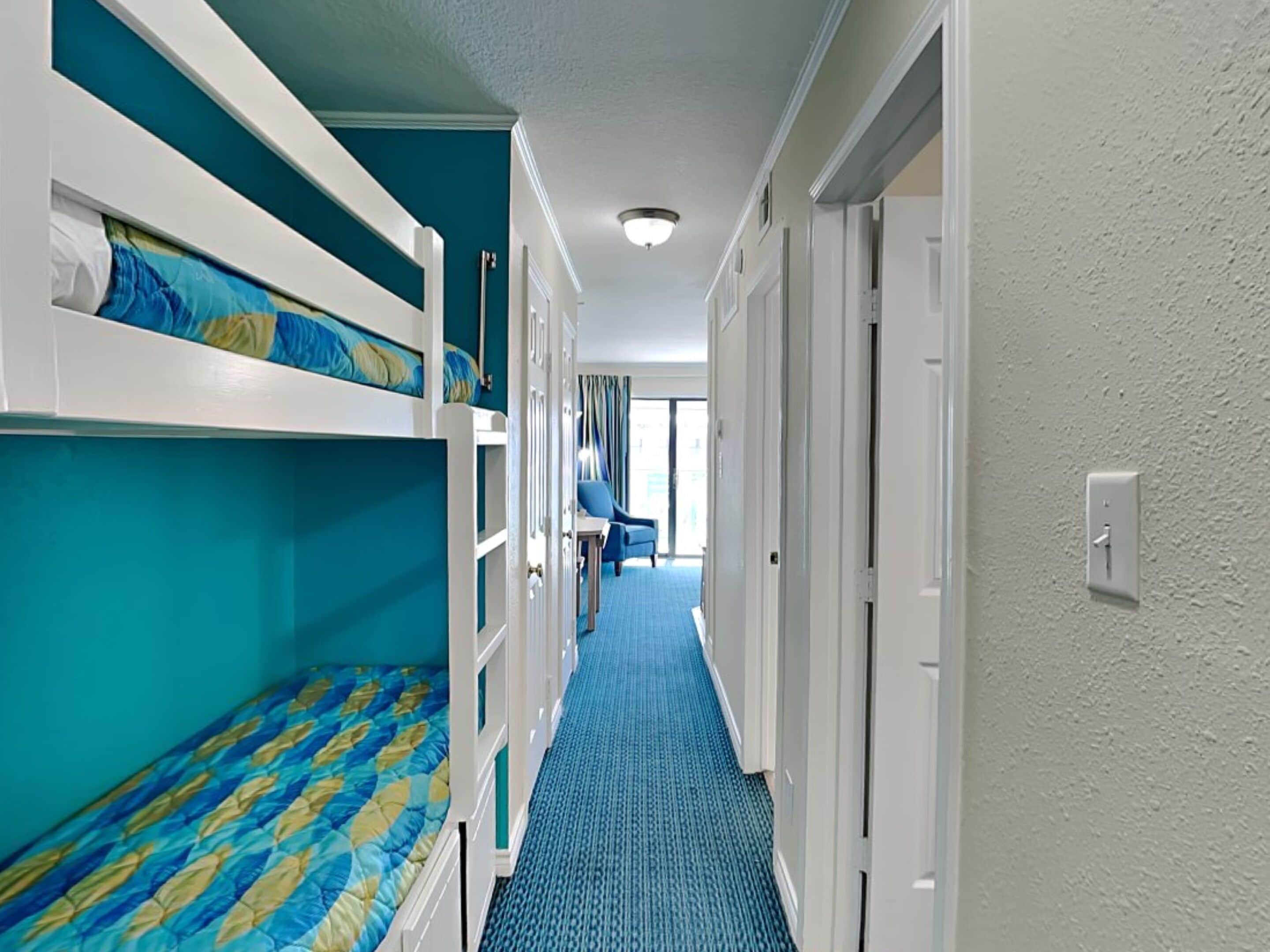Corredor com beliches de um lado e porta que dá para um quarto com carpete azul.