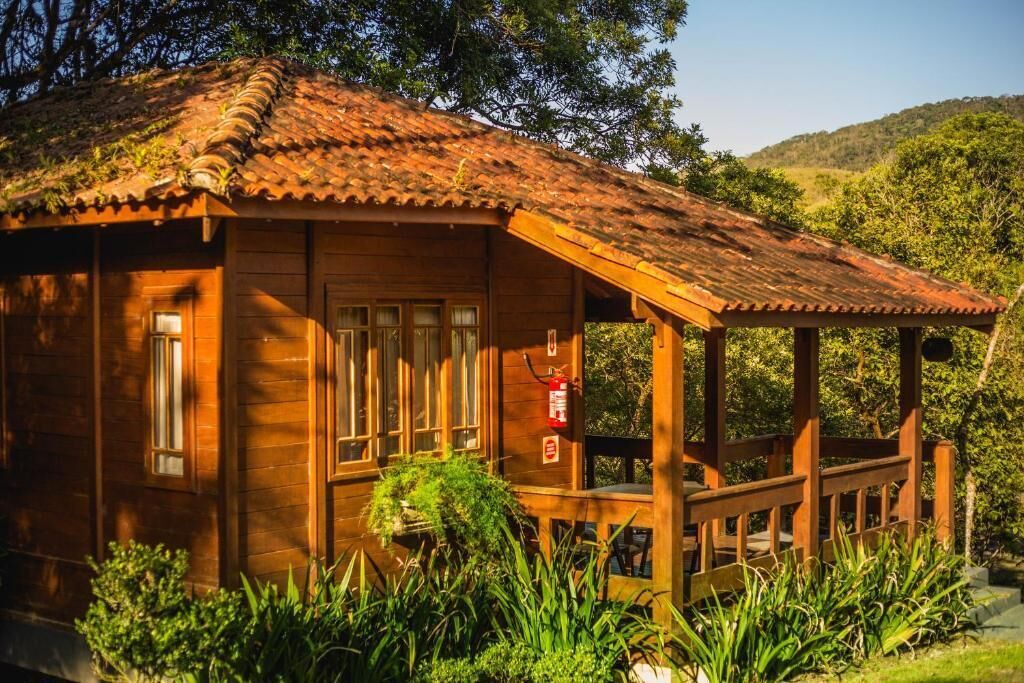 Uma cabana de madeira com telhado de telhas, rodeada por vegetação exuberante e colinas ao fundo.
