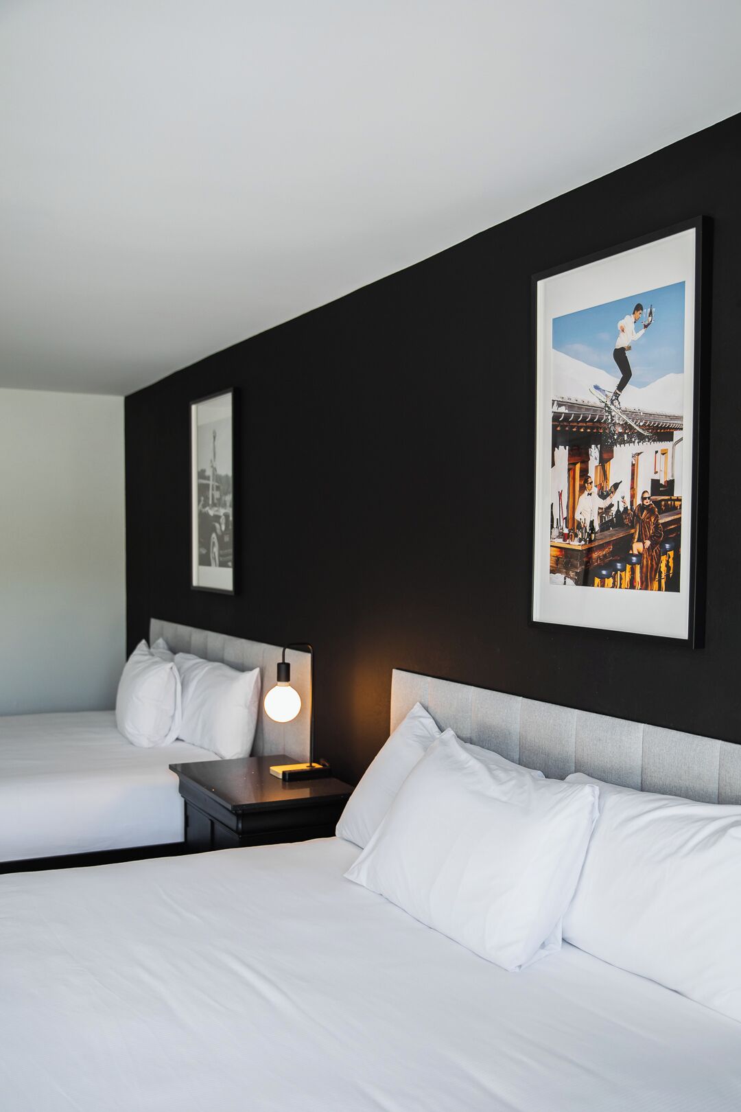 Quarto de hotel com duas camas, uma mesa de cabeceira com abajur e duas fotos emolduradas na parede.