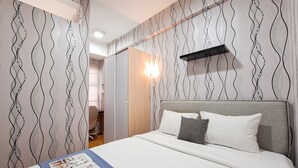 Cama bem arrumada com roupa de cama branca e cinza, um cobertor azul e uma revista ao lado. Cabeceira de madeira e prateleira fixada na parede são visíveis.