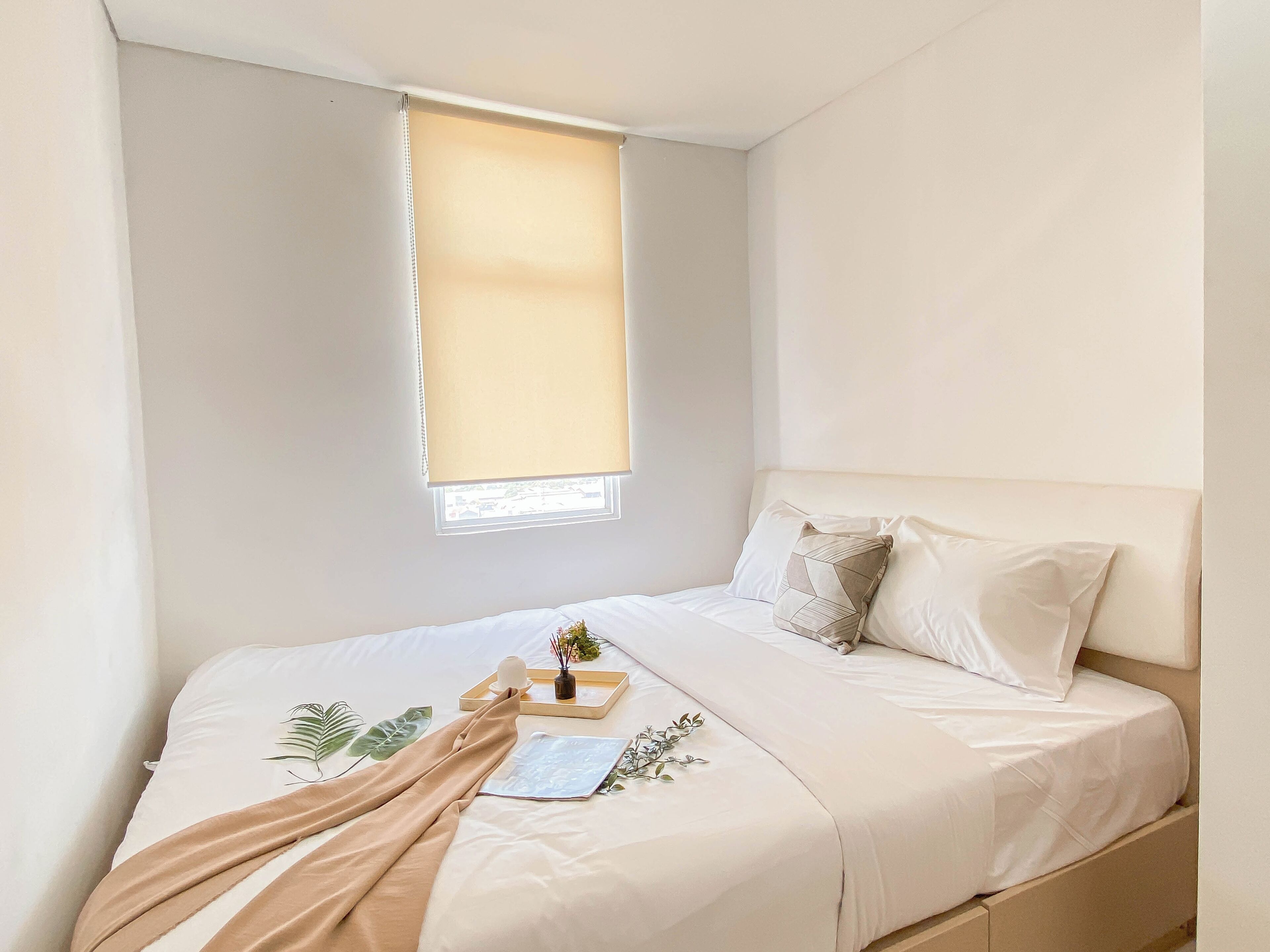 Quarto com cama, roupa de cama branca, janela com persiana e mesa de cabeceira com abajur e bandeja.