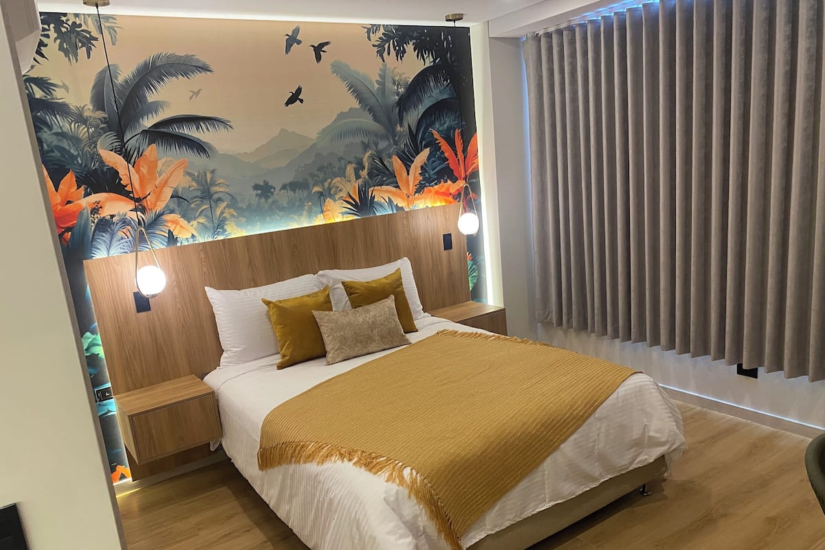 Quarto com cama, cabeceira de madeira e um mural com paisagem tropical.