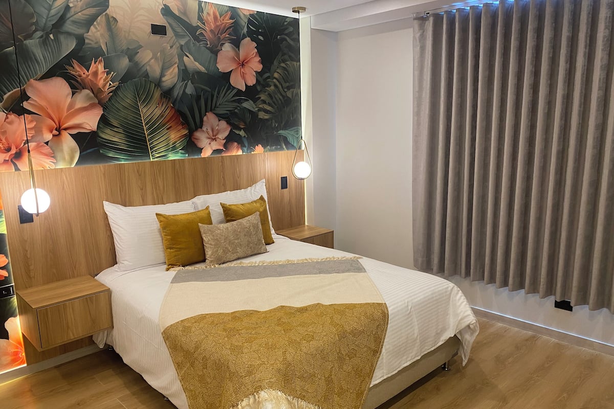 Quarto com cama, cabeceira de madeira, mesas de cabeceira, painel na parede e janela com cortinas.