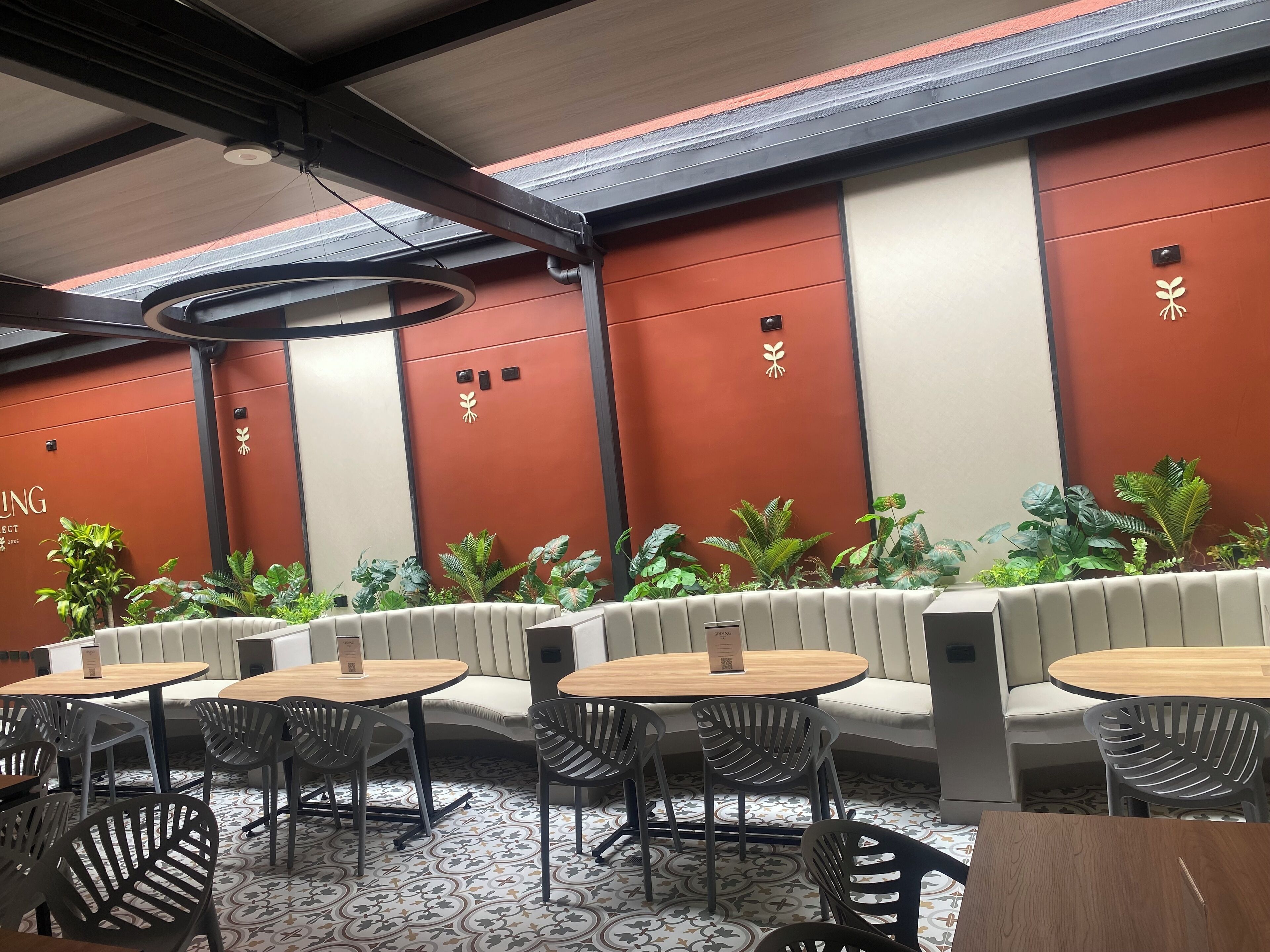 Interior de restaurante com mesas redondas, cadeiras pretas, um banco acolchoado e plantas em vasos.