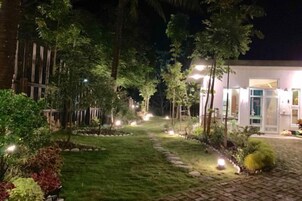 Uma passagem iluminada em um jardim noturno, levando a um edifício com porta de vidro, cercado por árvores e com iluminação no chão.